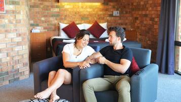 Um casal sentado em um sofá em um quarto com parede de tijolos e uma cama com travesseiros vermelhos.