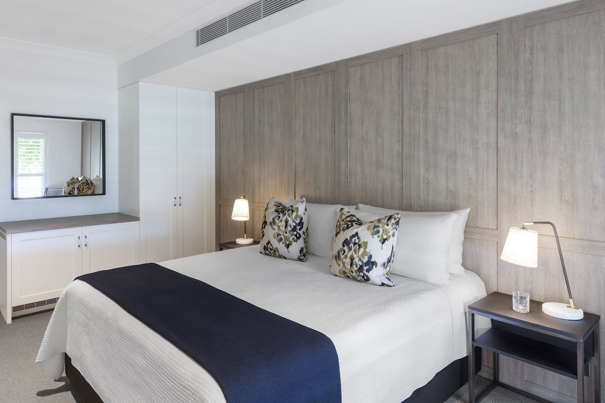 Quarto de hotel com uma cama grande, mesa de cabeceira com luminária, guarda-roupa e espelho na parede.