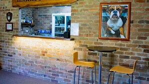 Paredes de tijolo com uma placa de madeira que diz “HARRY'S BAR” acima de um balcão com prateleiras cheias de garrafas. Há duas cadeiras amarelas e um quadro com a imagem de um cão na parede.