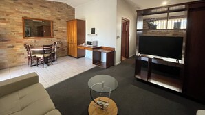 Sala de estar com televisão, área de jantar com cadeiras, cozinha equipada com micro-ondas e ventoinha no teto.