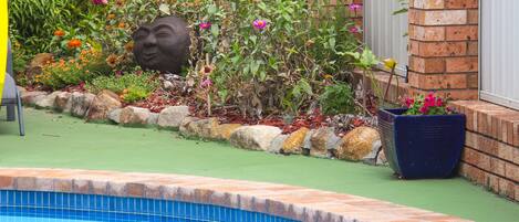 Piscina com borda azulejada, rodeada por um jardim com diversas plantas e um rosto de pedra decorativo.