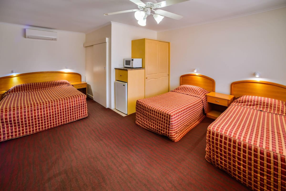 Quarto de hotel com duas camas de solteiro, um guarda-roupa de madeira, uma kitchenette pequena e um ventilador de teto.