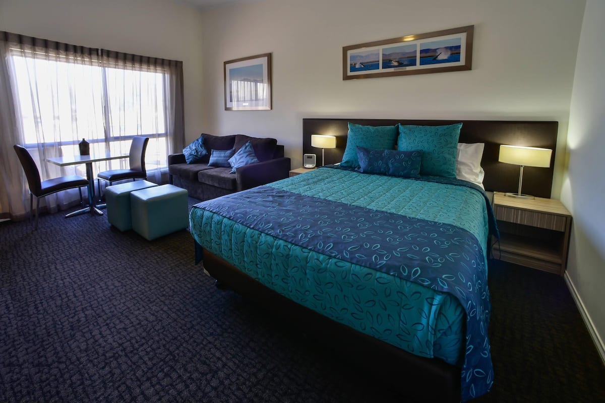 Quarto de hotel com uma cama grande, um sofá, uma escrivaninha e uma cadeira. Há duas mesas de cabeceira com abajures.