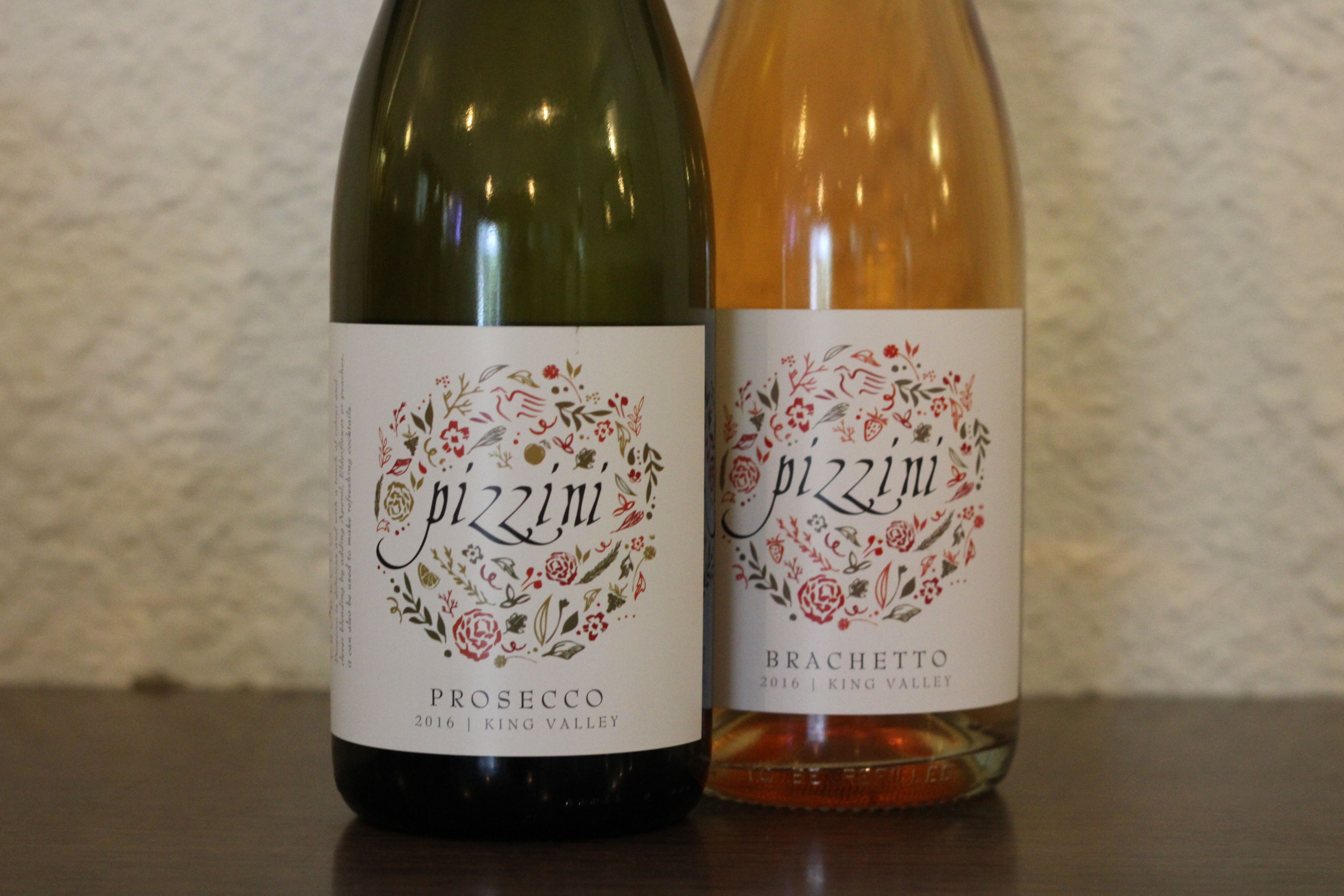 Duas garrafas de vinho, uma com o rótulo 'PROSECCO' e outra com 'BRACCHETTO', ambas da marca 'Pizzini' e da região 'King Valley'.