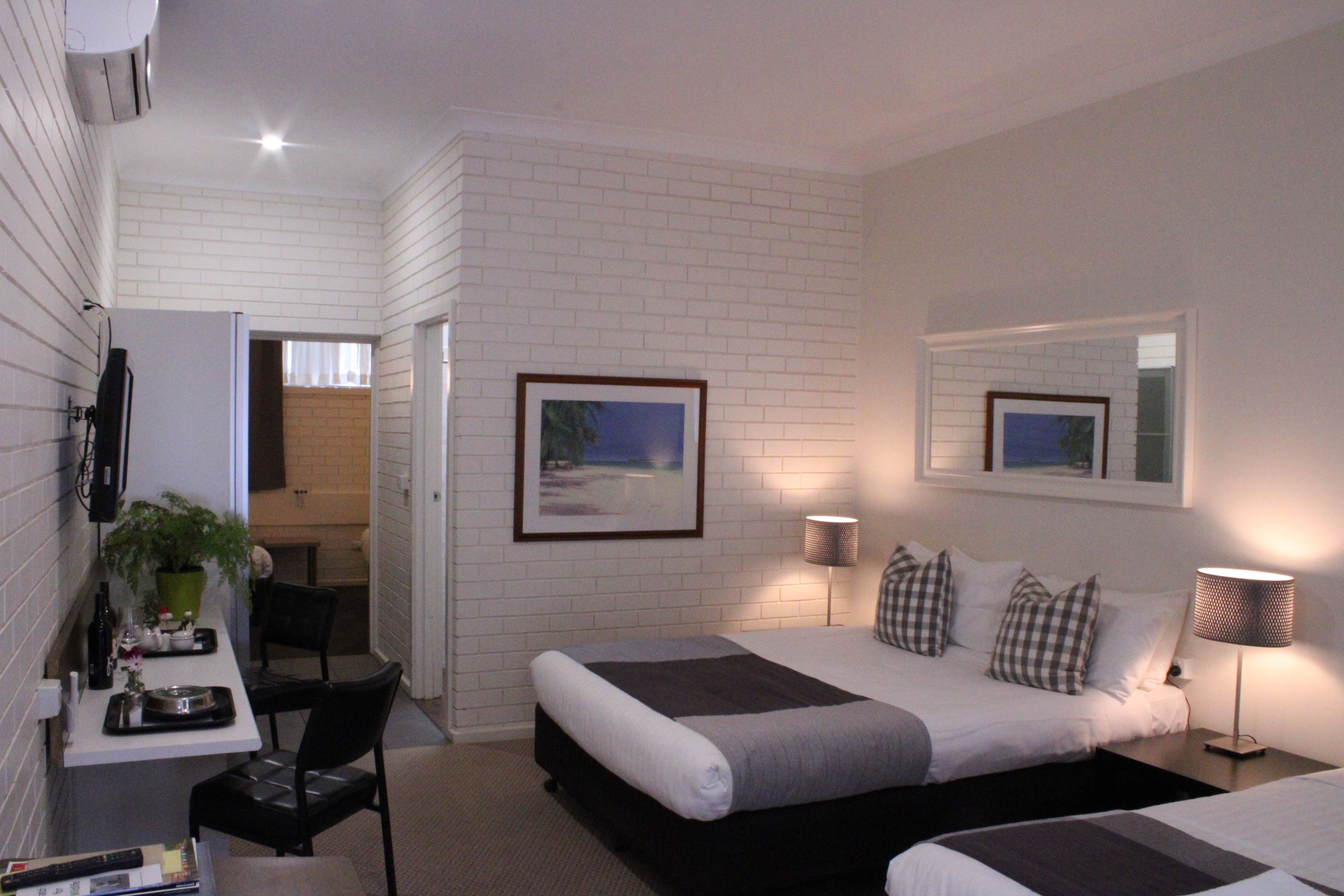 Quarto de hotel com parede de tijolos brancos, cama com colcha cinza e branca, mesa de cabeceira com abajur, uma cadeira e uma pequena escrivaninha com uma planta e um controle remoto.