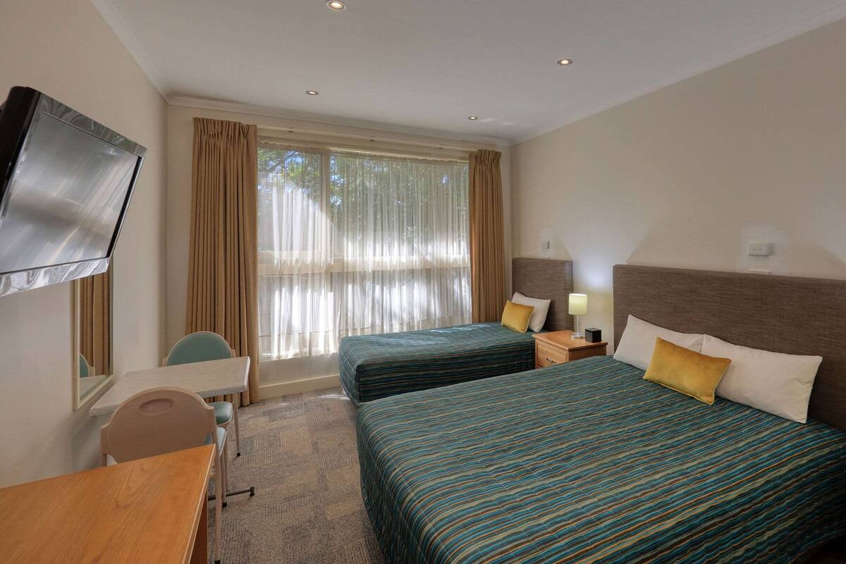 Quarto de hotel com duas camas, uma escrivaninha, uma cadeira, uma televisão e uma janela com cortinas.