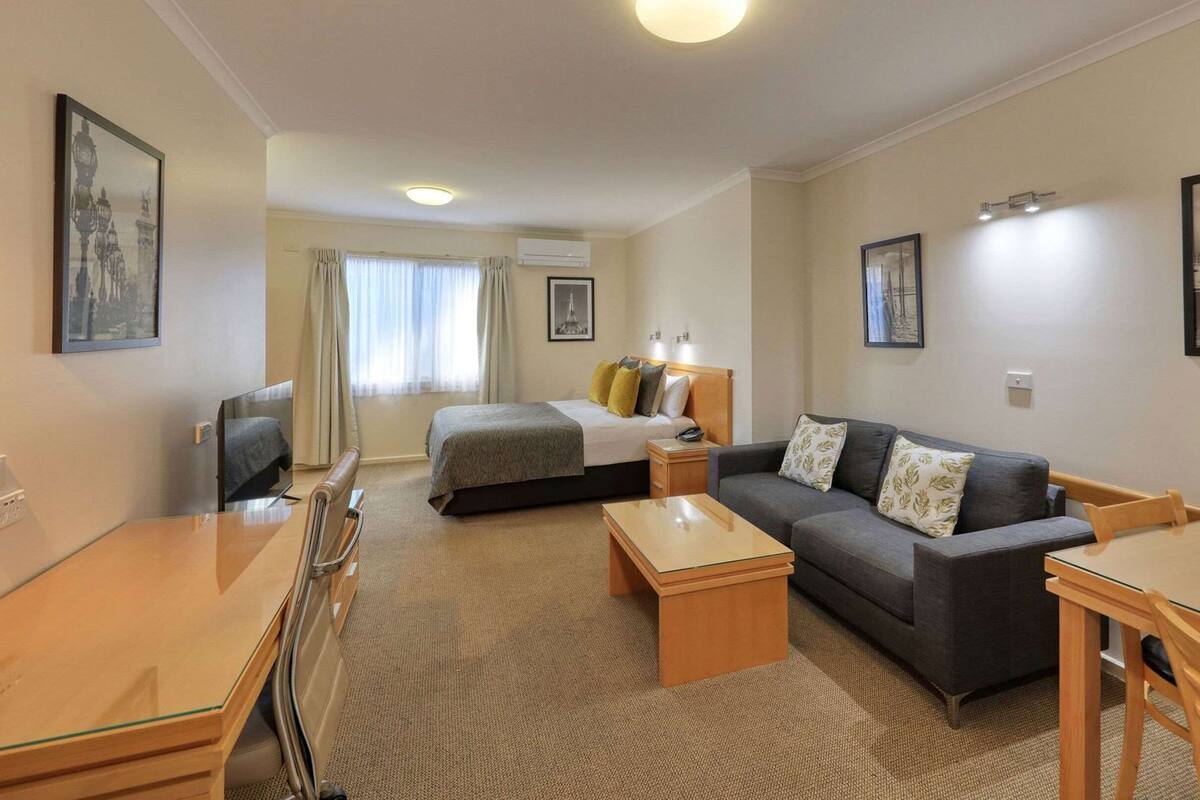 Quarto de hotel com cama, sofá, escrivaninha e cadeira. Há uma janela com cortinas, ar-condicionado fixado na parede e quadros decorativos.