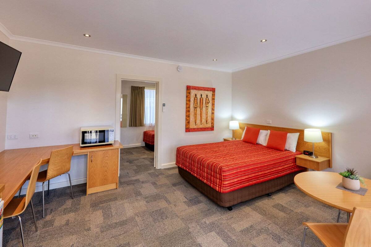 Quarto de hotel com cama, escrivaninha, cadeira e televisão na parede.