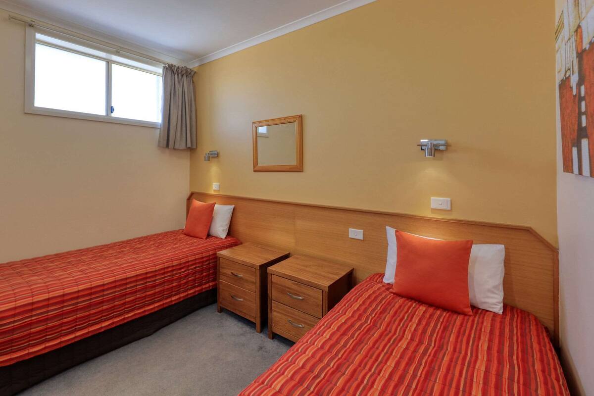 Quarto de hotel com duas camas de solteiro, uma pequena mesa de cabeceira de madeira, um espelho e uma janela com cortinas.