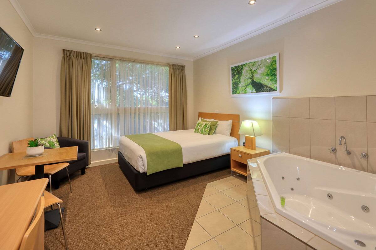Quarto de hotel com cama, escrivaninha, cadeira, janela grande com cortinas, televisão na parede e banheiro com banheira, além de uma planta verde na escrivaninha.