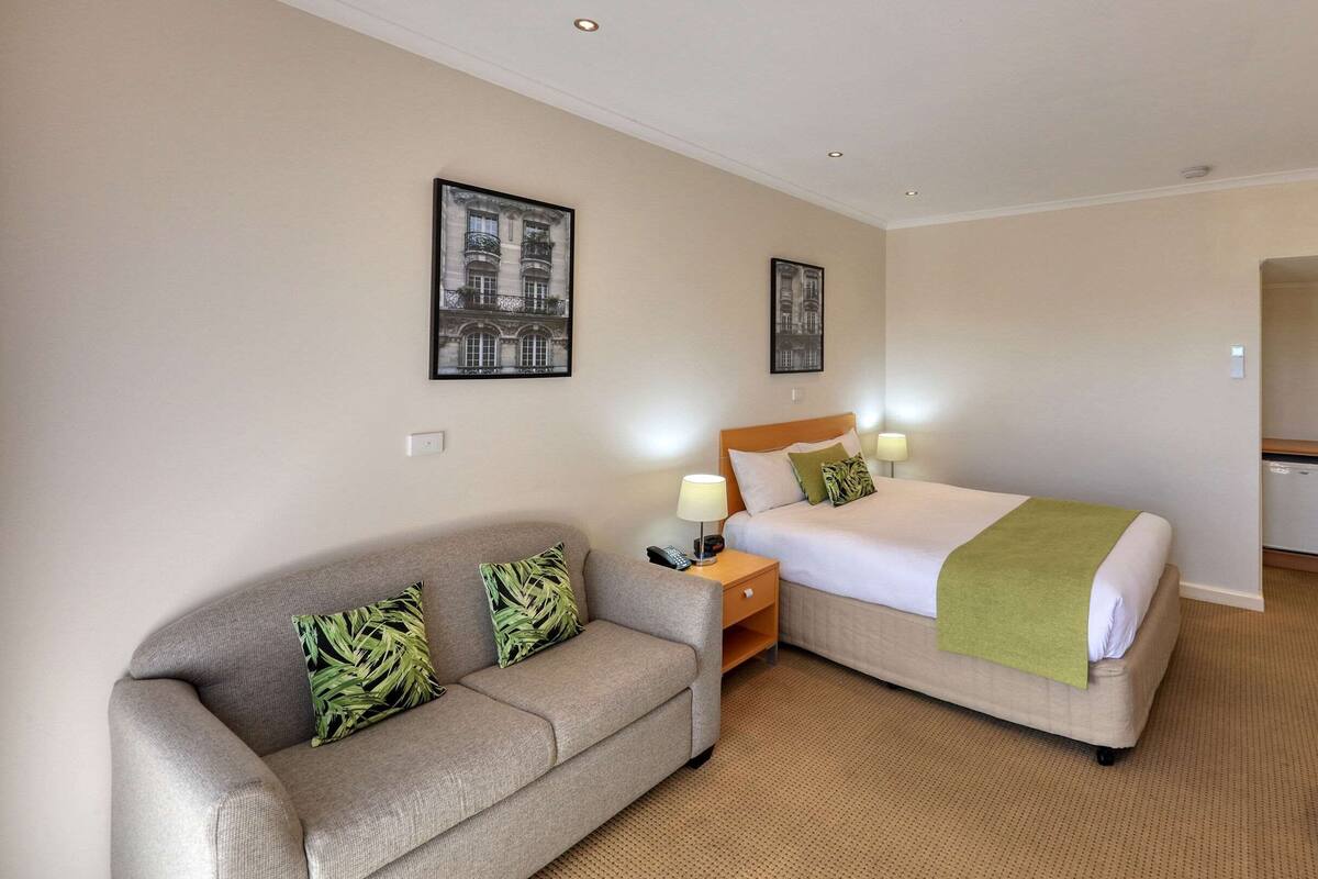 Quarto de hotel com cama, sofá, mesa de trabalho e cozinha compacta.