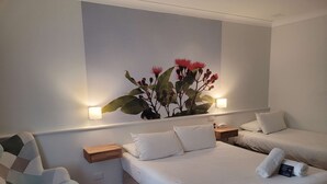Quarto de hotel com duas camas, um sofá, uma planta na parede e luminárias de cabeceira.