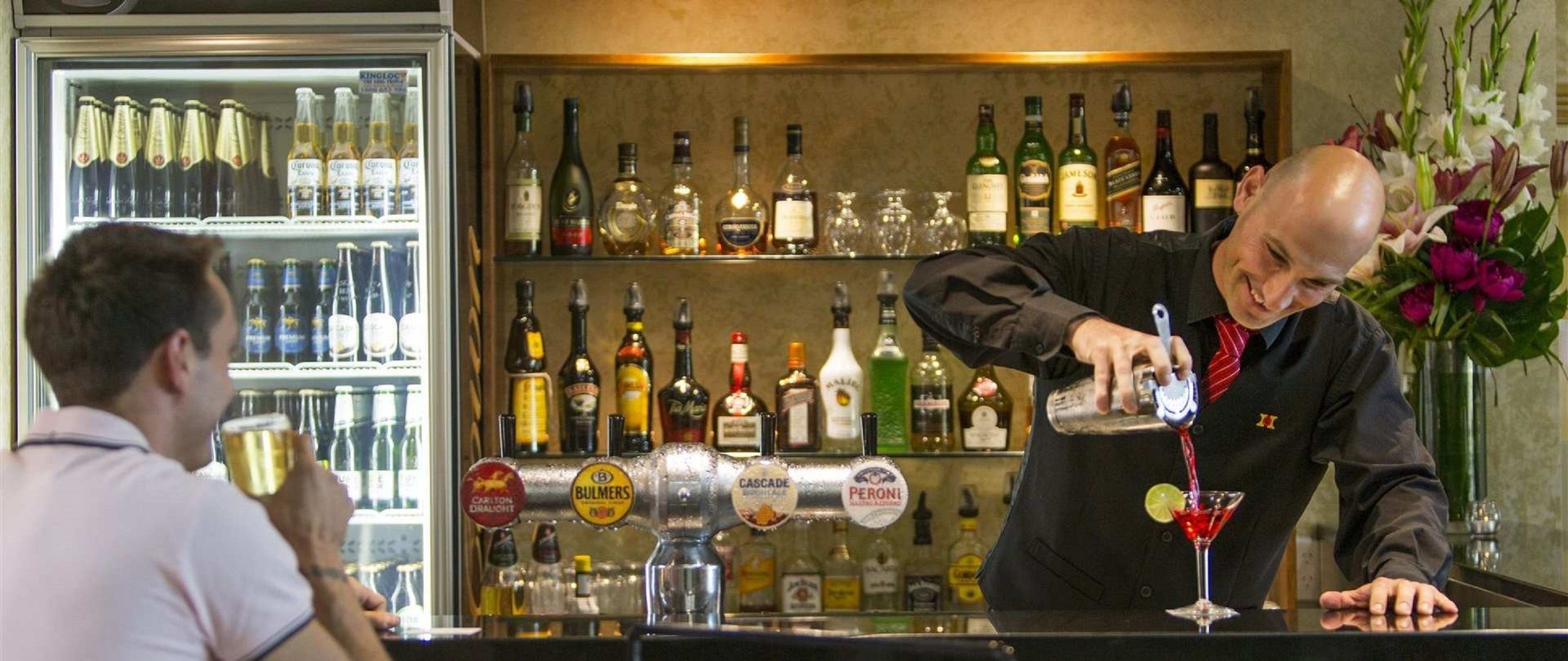 Uma área de bar com um bartender servindo uma bebida em taça de martini, prateleiras com diversas garrafas de licor e uma vitrine refrigerada.