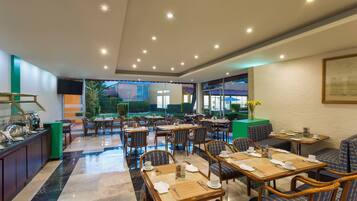 Uma sala de jantar de hotel com várias mesas e cadeiras, um balcão de buffet e vista para o exterior.