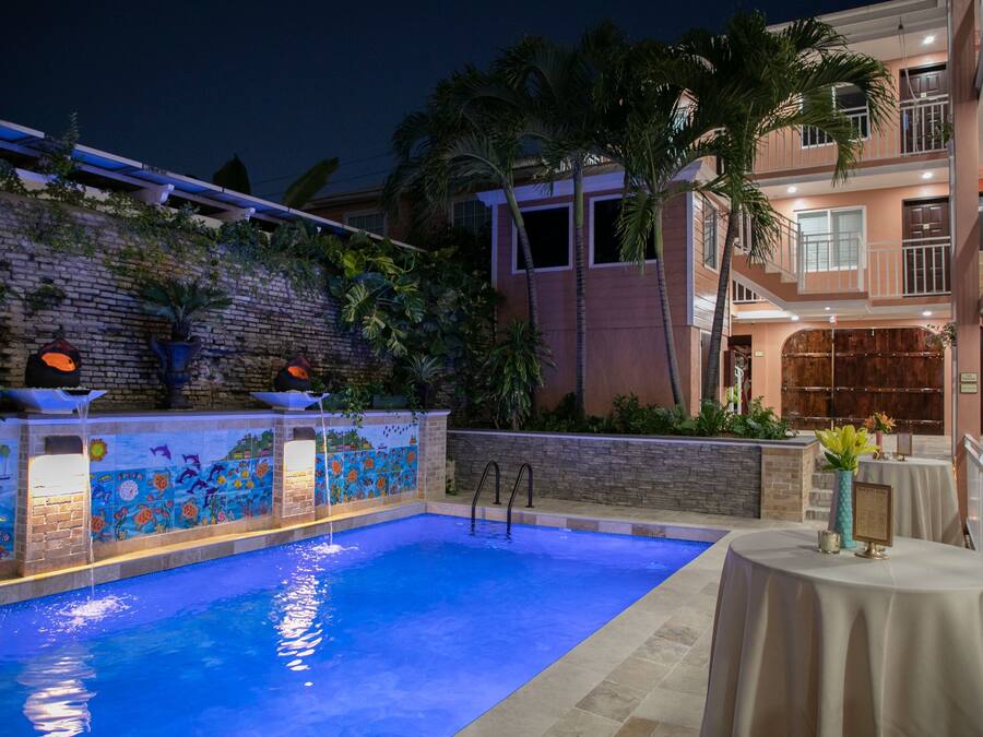 Piscina com borda azulejada e parede decorativa, rodeada por palmeiras e um edifício com varandas.