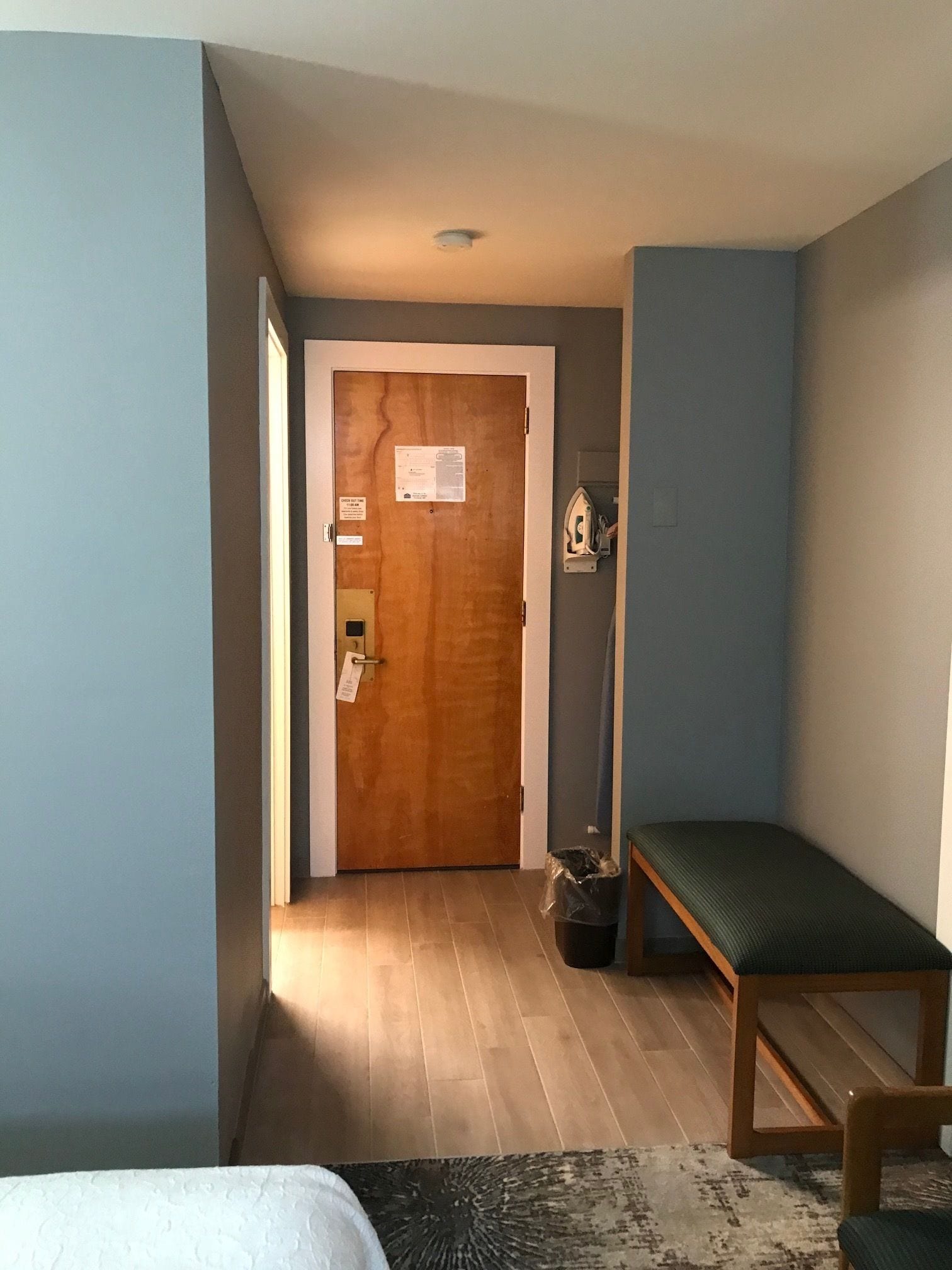 Um quarto com porta de madeira, um banco, uma lixeira e uma pequena mesa.