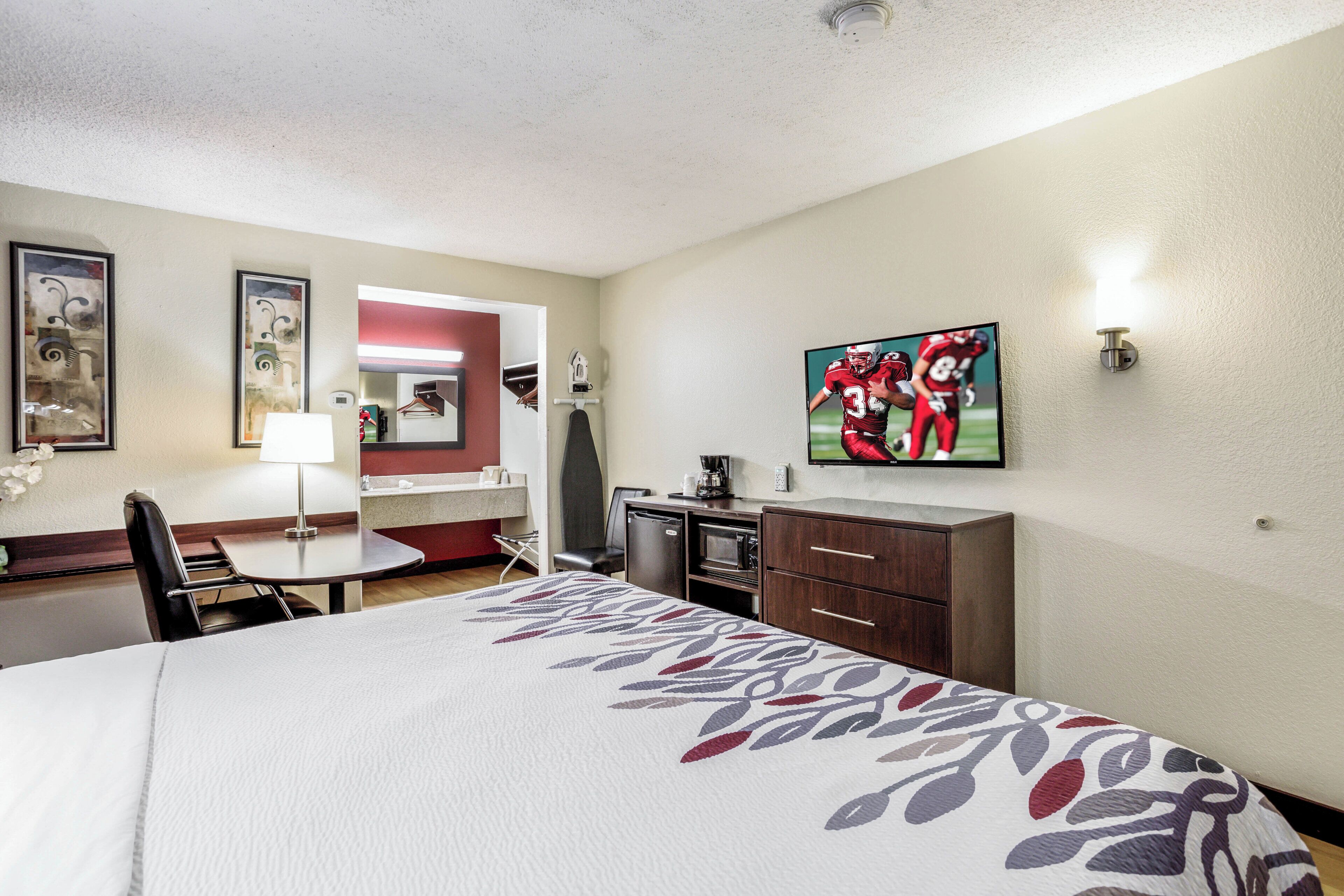 Quarto de hotel com cama, escrivaninha, cadeira, televisão e um quadro na parede.