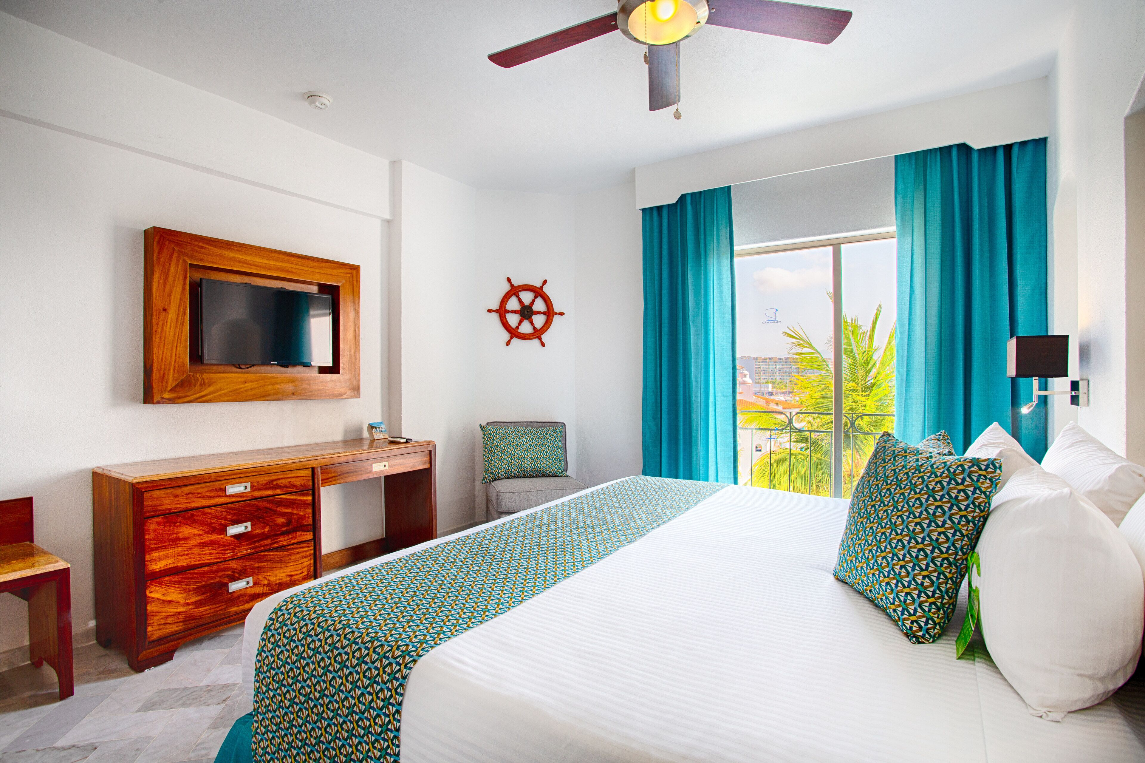 Quarto de hotel com cama, escrivaninha, cadeira, televisão, ventoinha no teto e vista para o exterior.