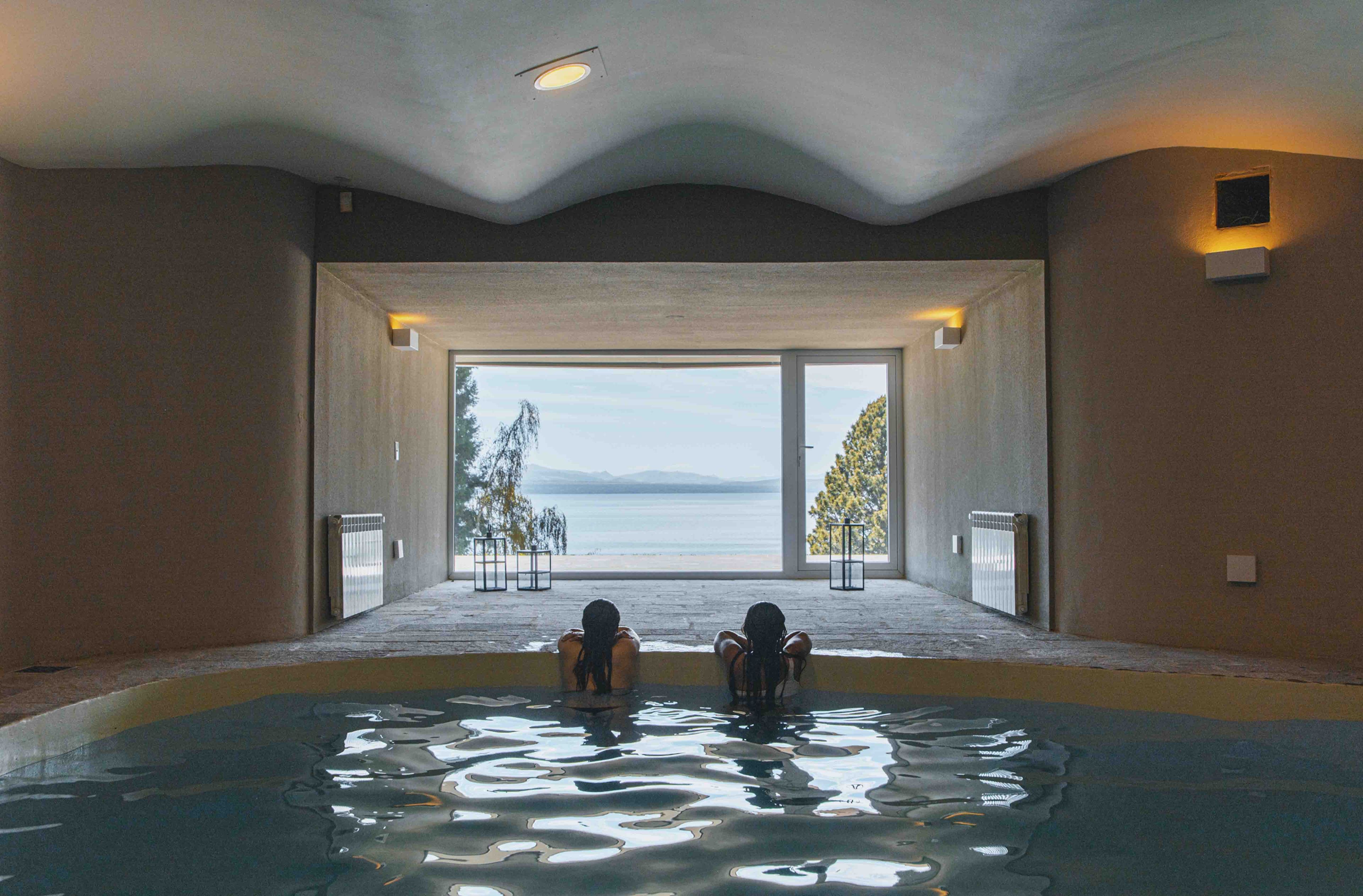 Piscina interior com duas pessoas na água, vista para o mar através de amplas janelas e teto com iluminação embutida.