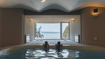Piscina interior com duas pessoas na água, vista para o mar através de amplas janelas e teto com iluminação embutida.