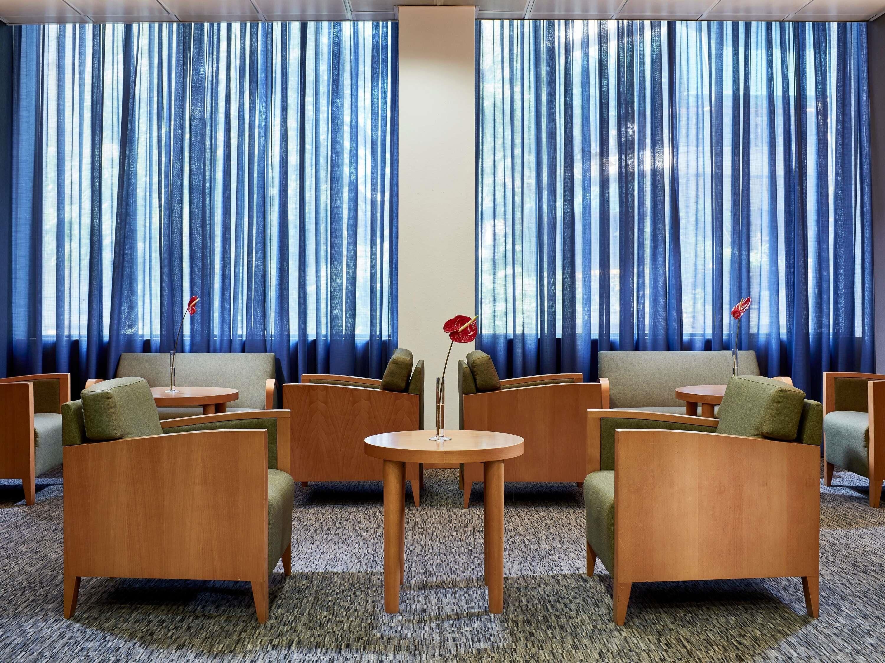 Um saguão de hotel com área de estar, janelas amplas com cortinas azuis e um vaso decorativo central.
