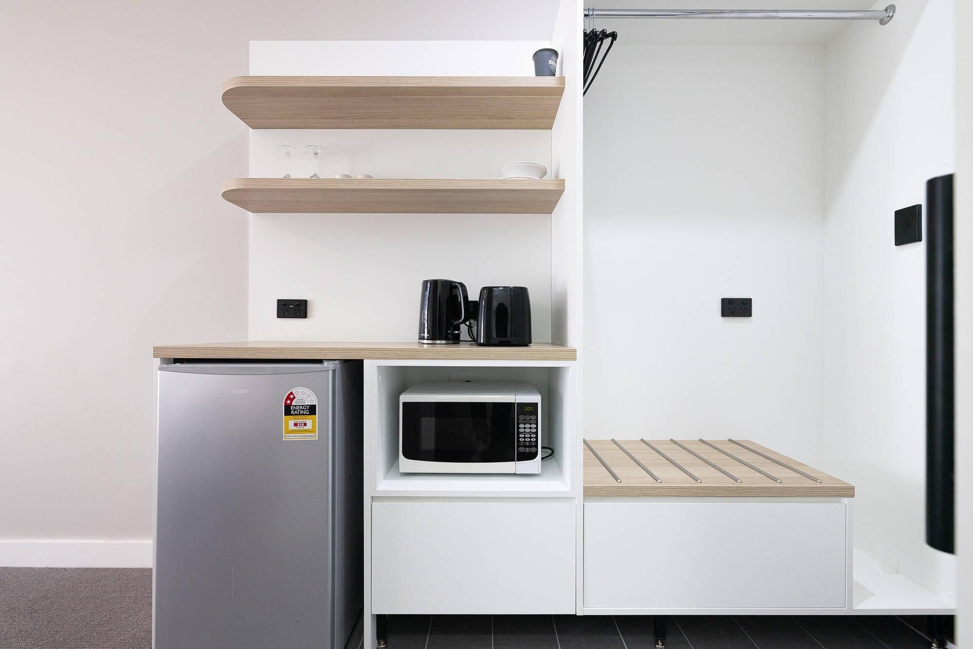 Uma kitchenette compacta com geladeira, micro-ondas, cafeteira e prateleiras de madeira.
