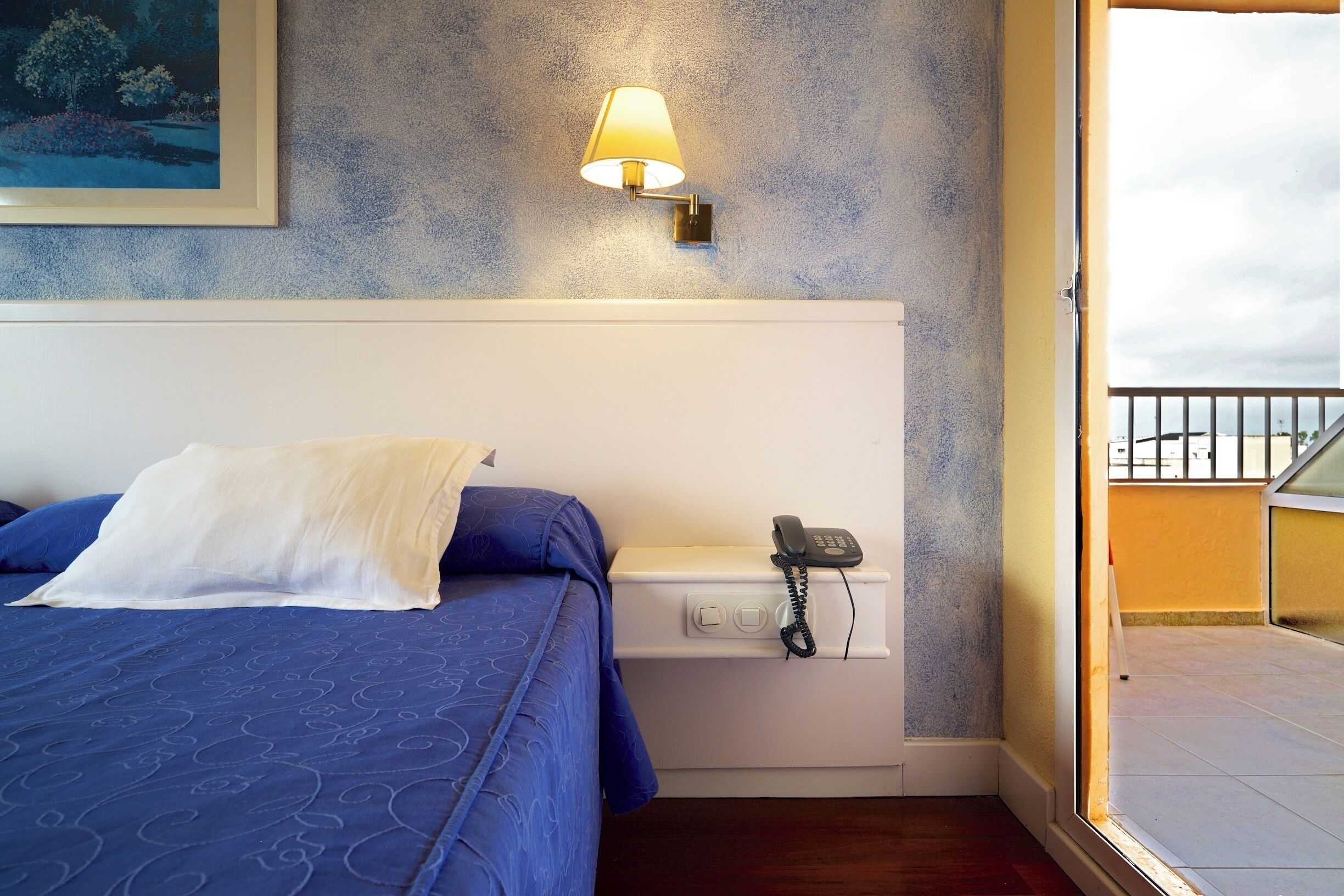 Quarto de hotel com cama, mesa de cabeceira com abajur, telefone e vista para a varanda.