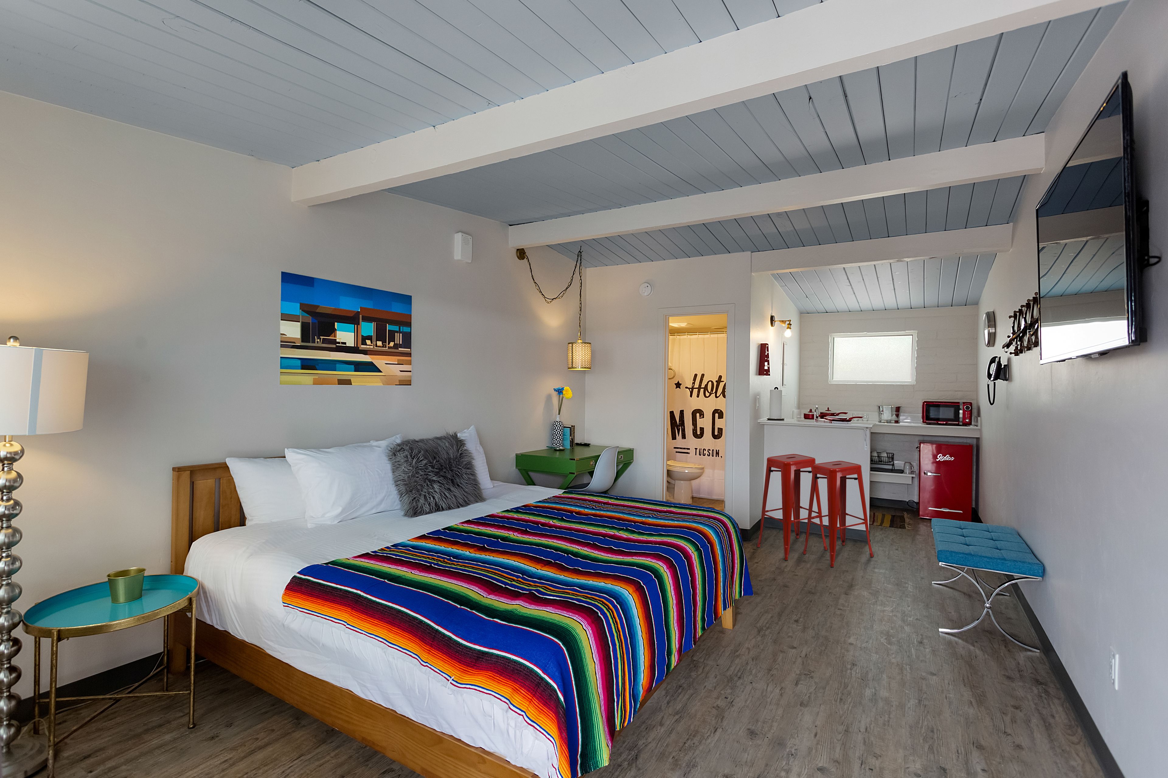 Quarto de hotel com cama, escrivaninha, cadeira, mesa pequena, abajur e televisão.