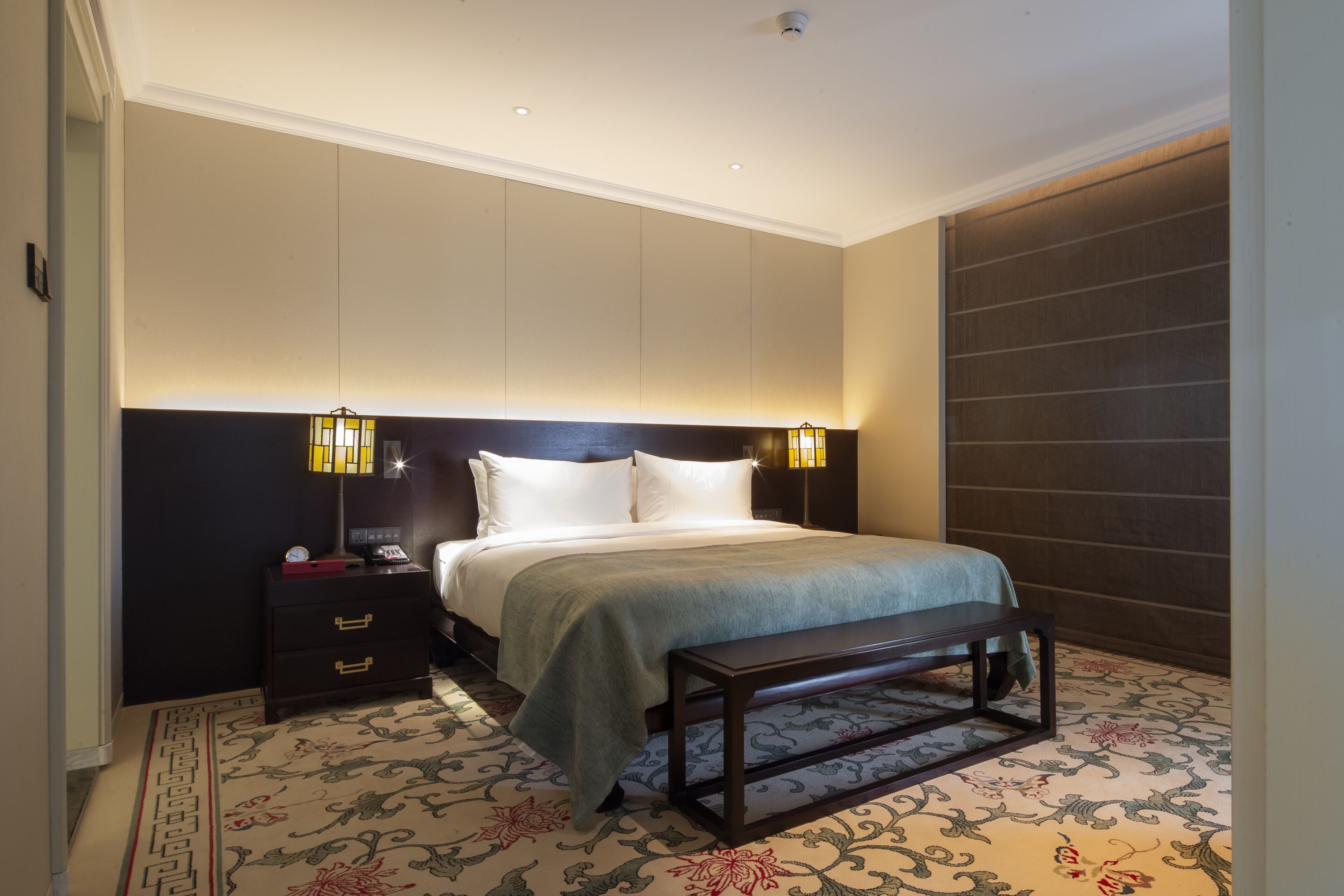 Quarto de hotel com cama, mesas de cabeceira, um banquinho e carpete estampado.