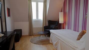 Quarto de hotel com cama, escrivaninha, cadeira, mesinha e janela com cortinas.
