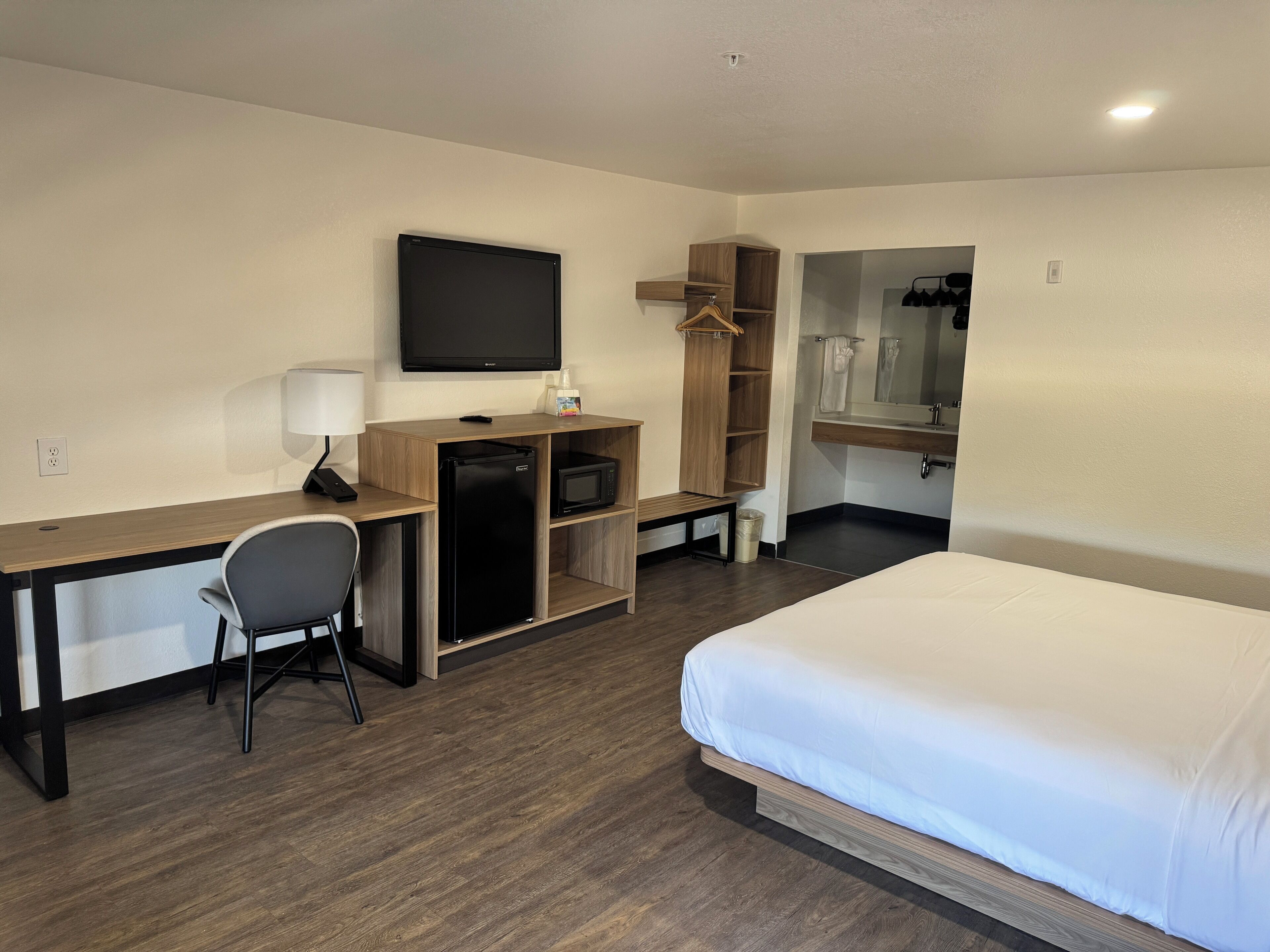 Quarto de hotel com cama, escrivaninha, cadeira, televisão, mini-geladeira, micro-ondas e banheiro com espelho e pia.