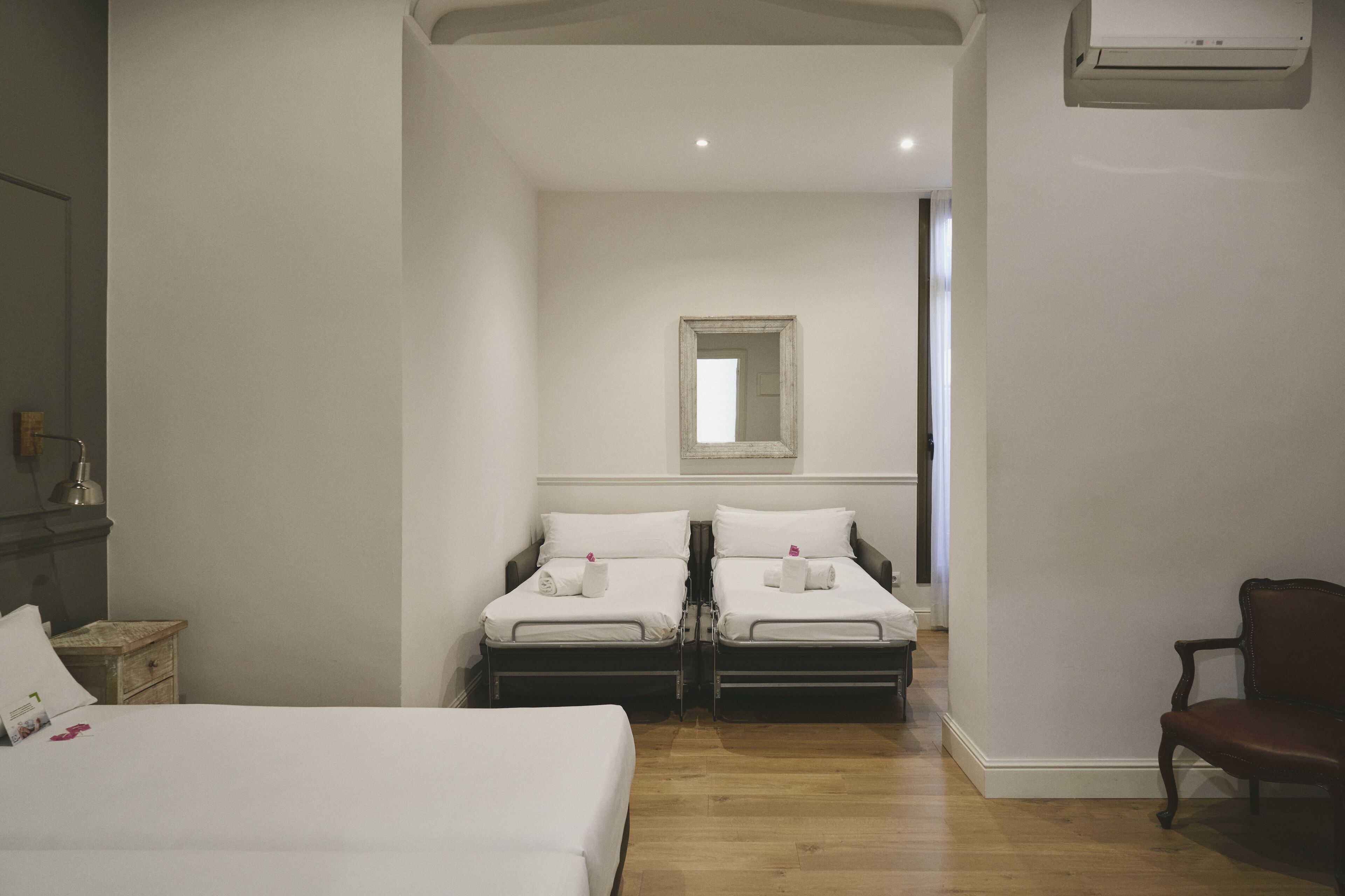 Quarto de hotel com duas camas, piso de madeira, uma cadeira e um espelho na parede.