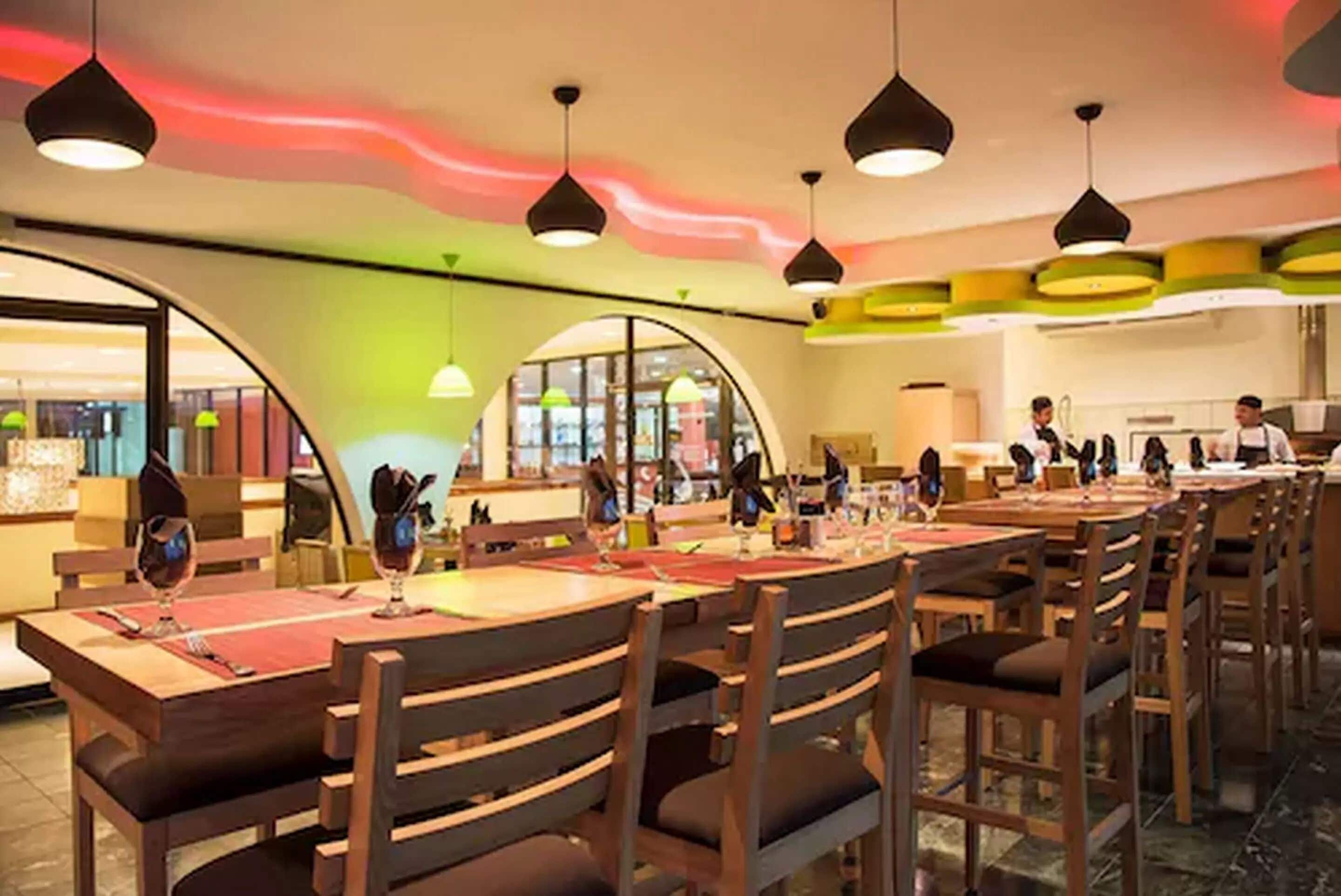 Um restaurante com mesas e cadeiras de madeira, iluminação pendente e vista para a cozinha.