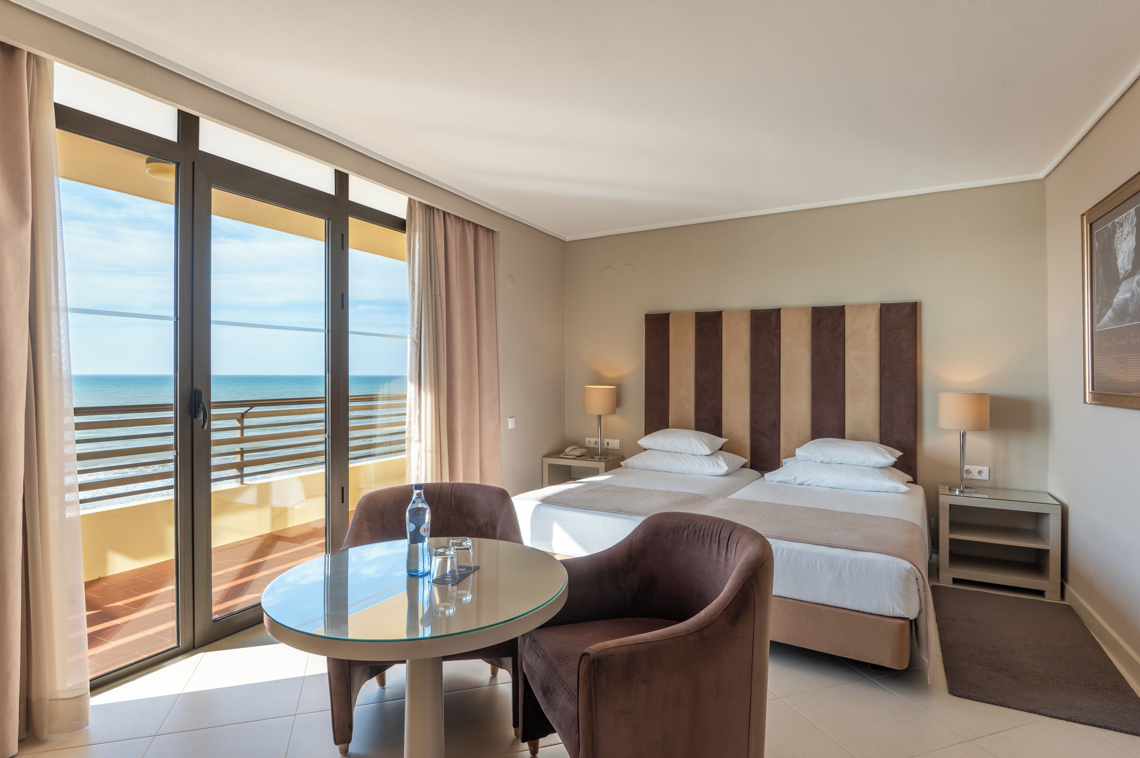 Quarto de hotel com cama, mesa de vidro, cadeira, mesinha de cabeceira e vista para o mar.