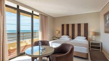 Quarto de hotel com cama, mesa de vidro, cadeira, mesinha de cabeceira e vista para o mar.