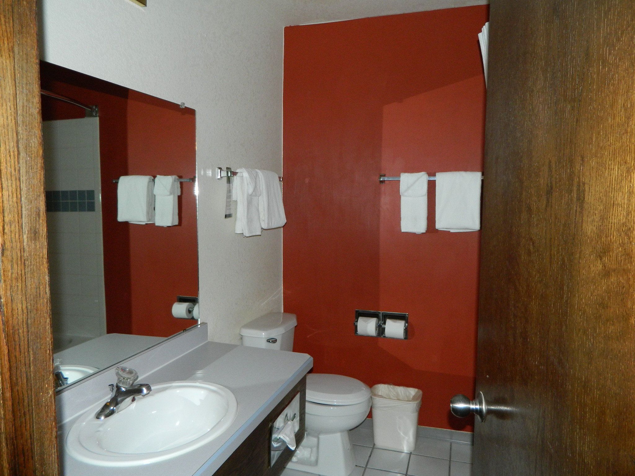 Casa de banho com lavatório branco, sanita e paredes vermelhas. Existem várias toalhas brancas penduradas na parede.