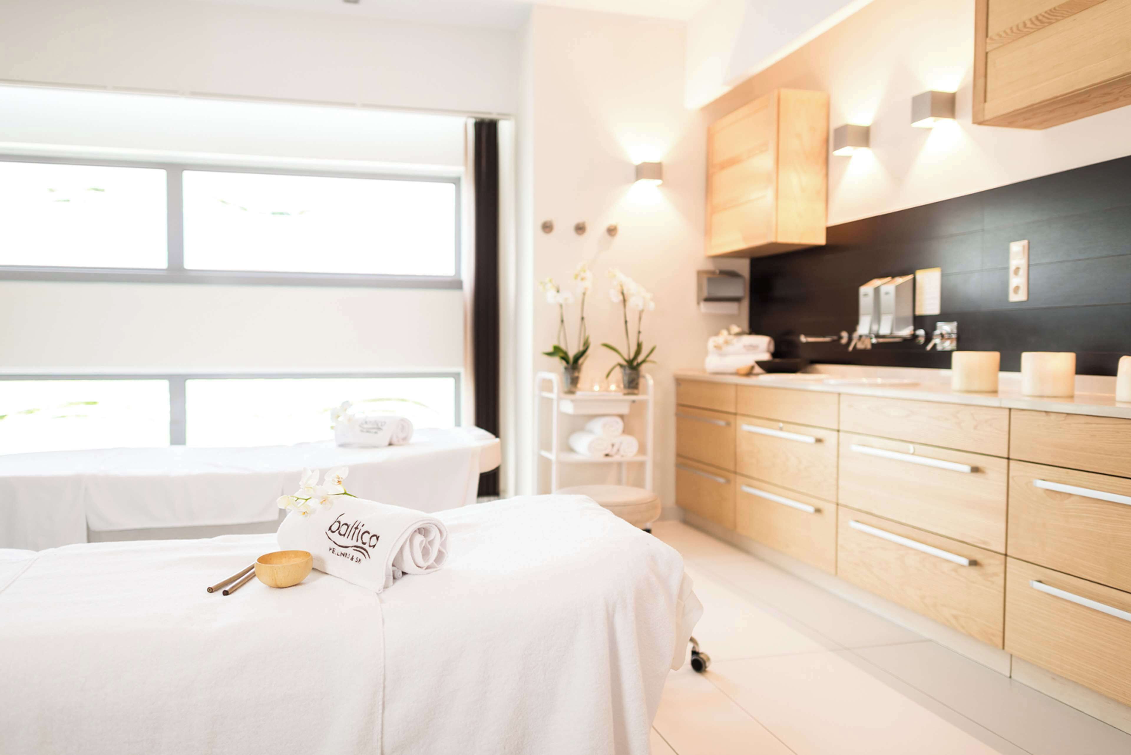 Quarto de hotel moderno com uma janela ampla, cama com colcha branca, pequena mesa com toalha e tigela de madeira, e uma kitchenette com armários de madeira e painel de fundo preto.
