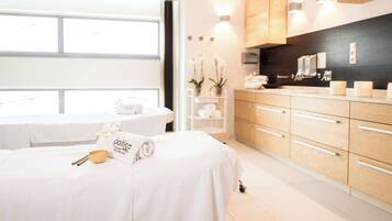 Quarto de hotel moderno com uma janela ampla, cama com colcha branca, pequena mesa com toalha e tigela de madeira, e uma kitchenette com armários de madeira e painel de fundo preto.