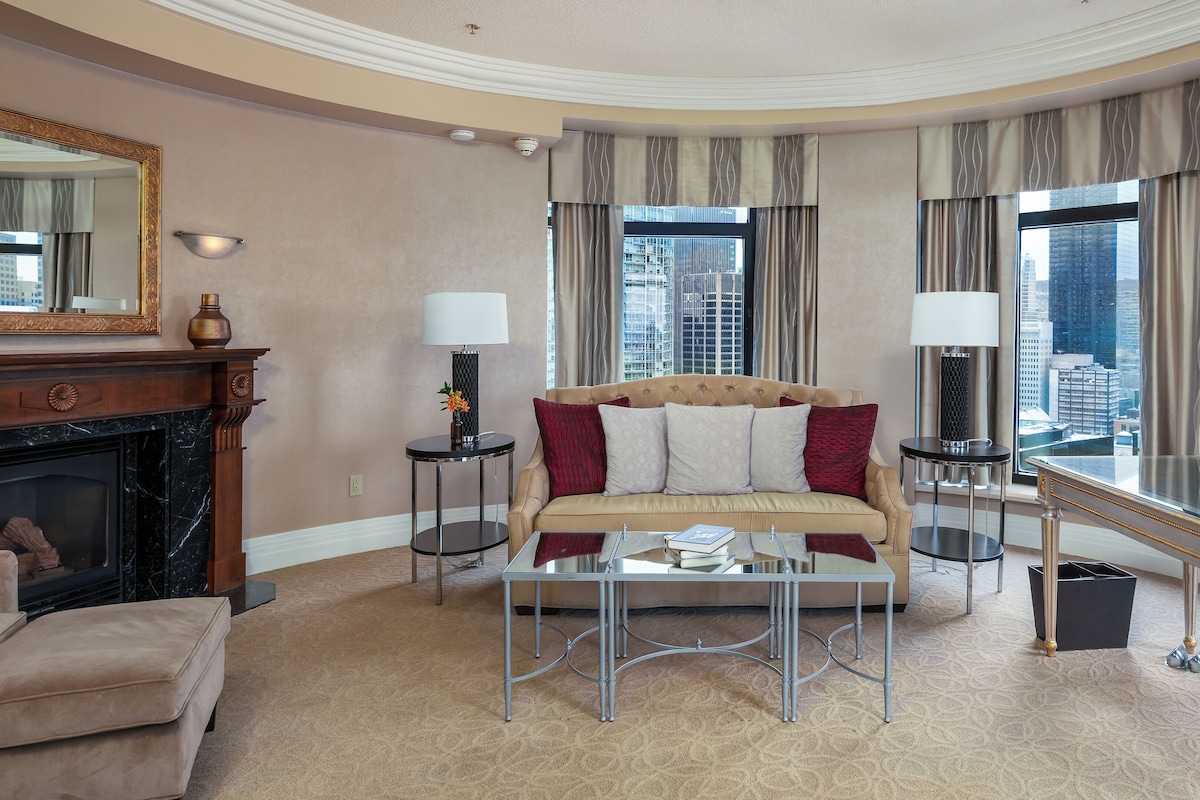 Quarto de hotel com sofá, duas mesas laterais, mesa de centro, lareira, espelho e vista para a cidade.