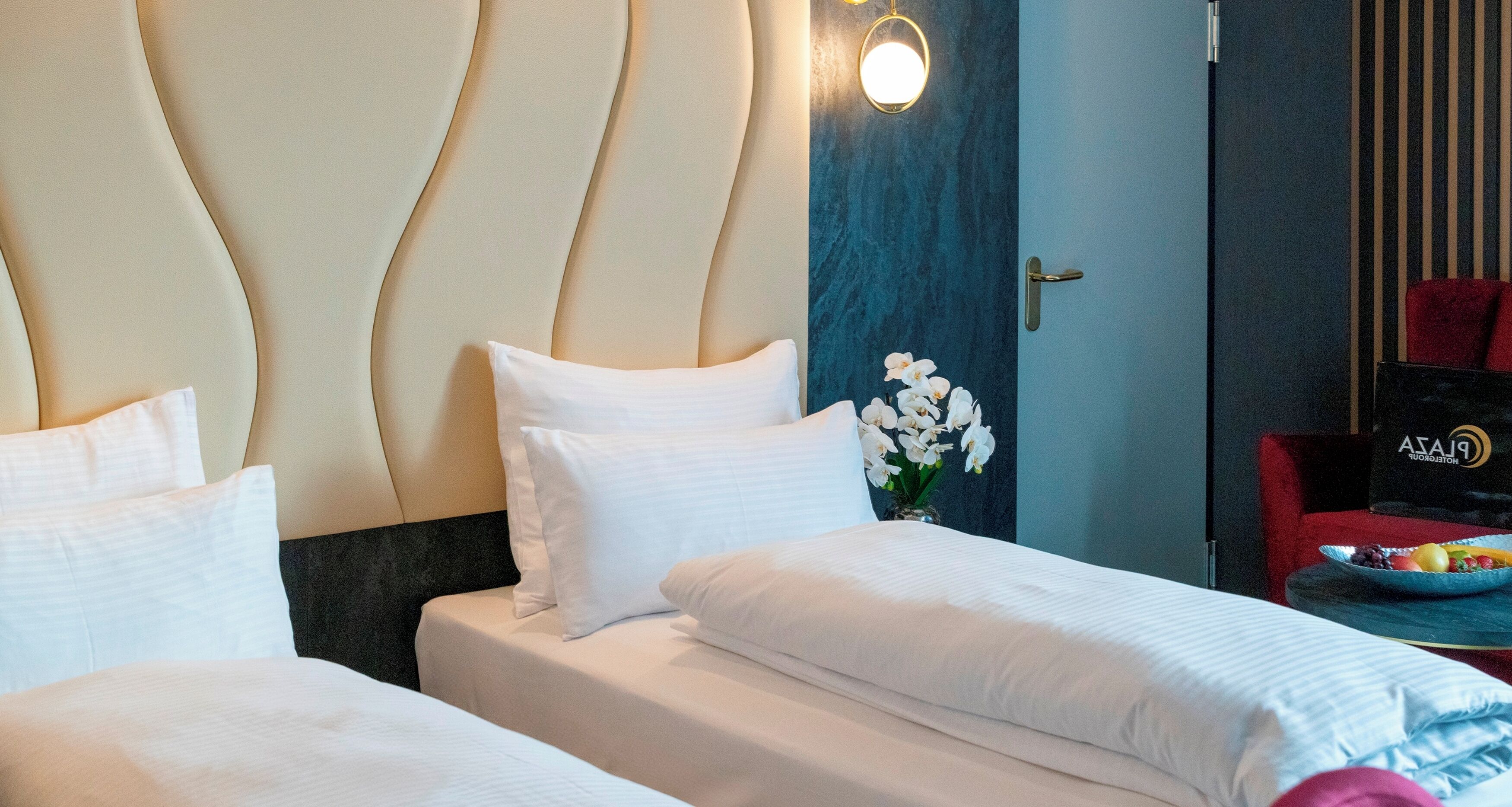 Quarto de hotel com duas camas, uma cabeceira moderna, um vaso com flores brancas, uma cadeira vermelha e um tabuleiro de café da manhã em uma mesa.