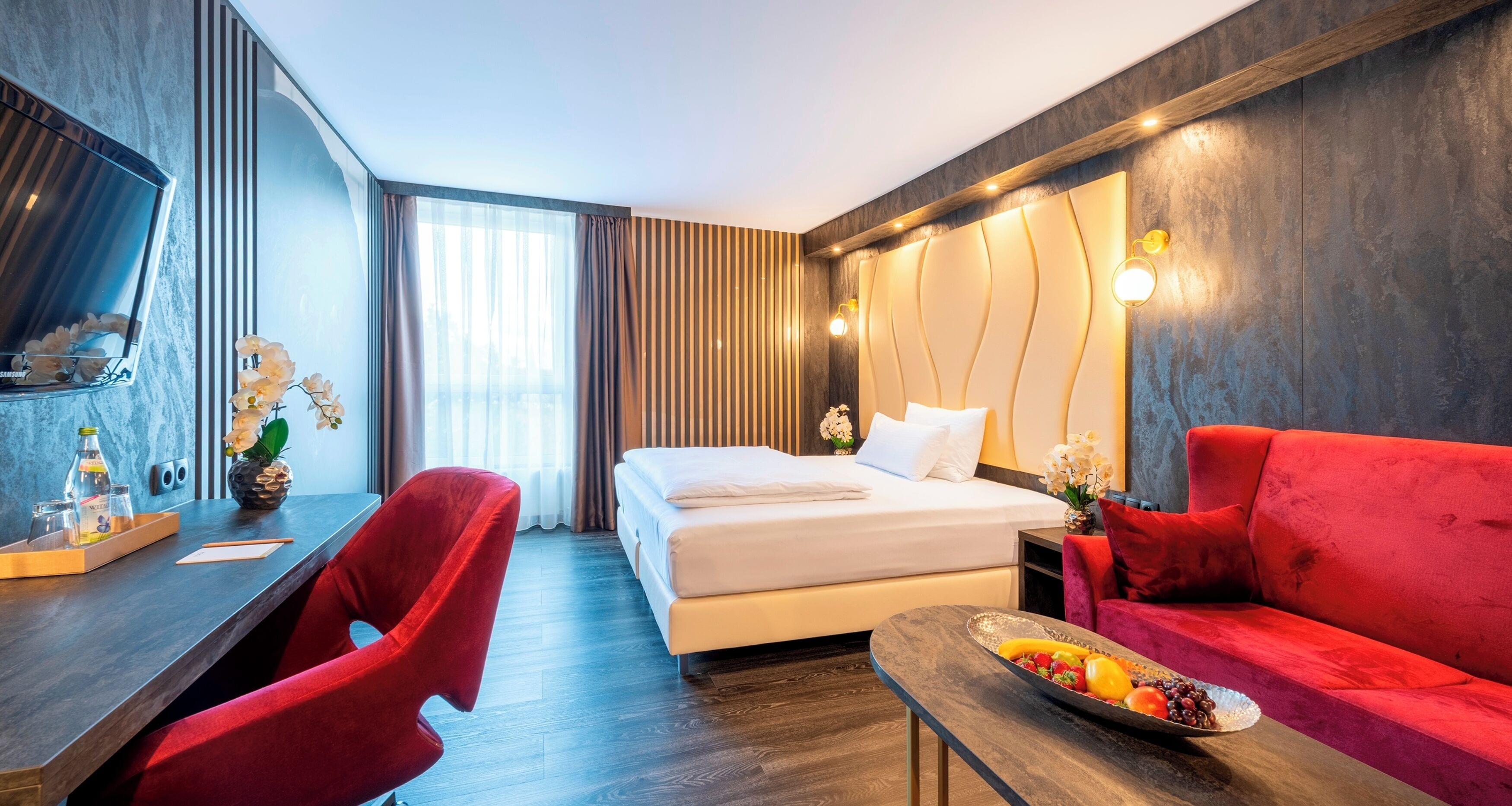 Quarto de hotel com uma cama, um sofá vermelho, uma escrivaninha com cadeira e uma televisão.