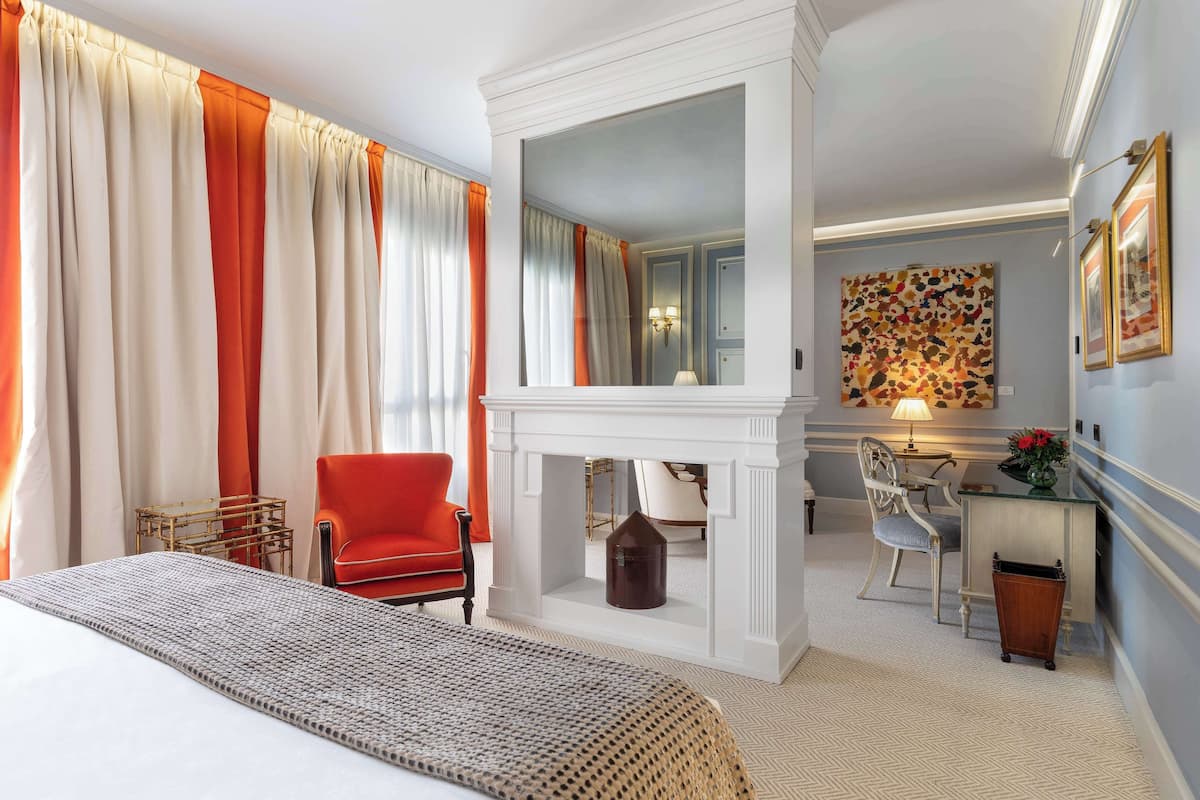 Quarto de hotel com cama, uma cadeira vermelha, escrivaninha com luminária, lareira e um espelho grande.