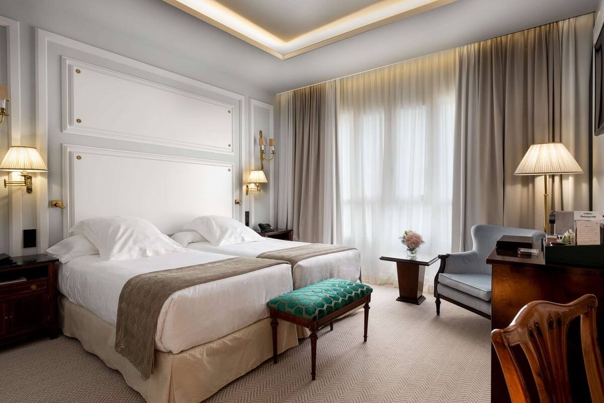 Quarto de hotel com duas camas, uma escrivaninha, uma cadeira e uma mesa pequena com abajur.