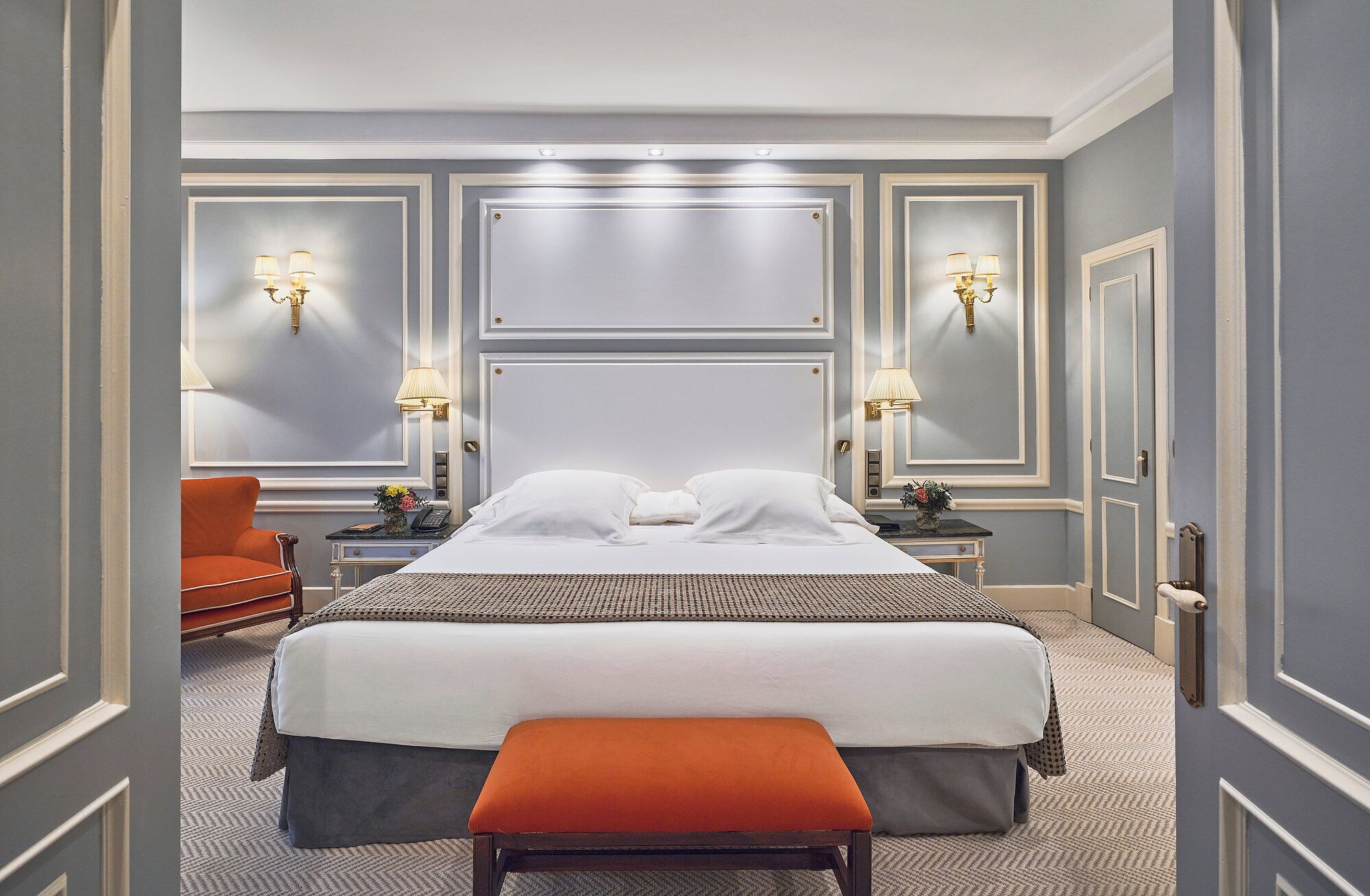 Quarto de hotel com cama grande, duas mesas de cabeceira com luminárias, uma cadeira e um banco.