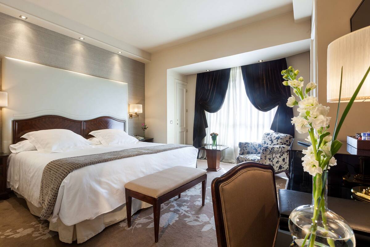 Quarto de hotel com uma cama grande, mesa de cabeceira, cadeira, escrivaninha e um vaso com flores.