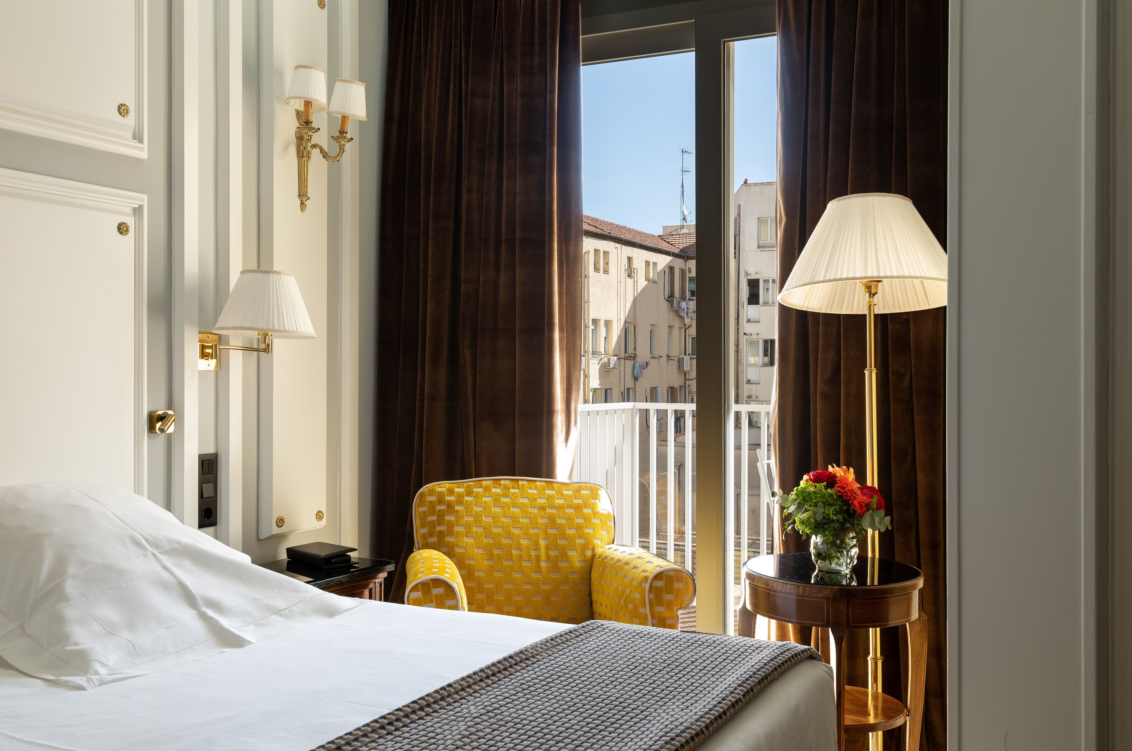 Quarto de hotel com cama, poltrona amarela, mesa pequena com um vaso de flores e uma luminária.