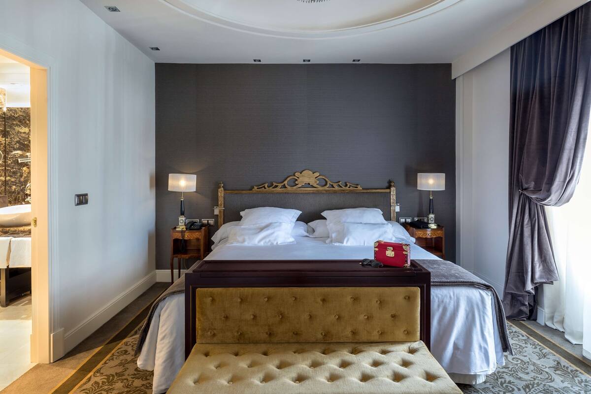 Quarto de hotel com uma cama grande, duas mesas de cabeceira, um criado-mudo, um banco e vista para um banheiro.