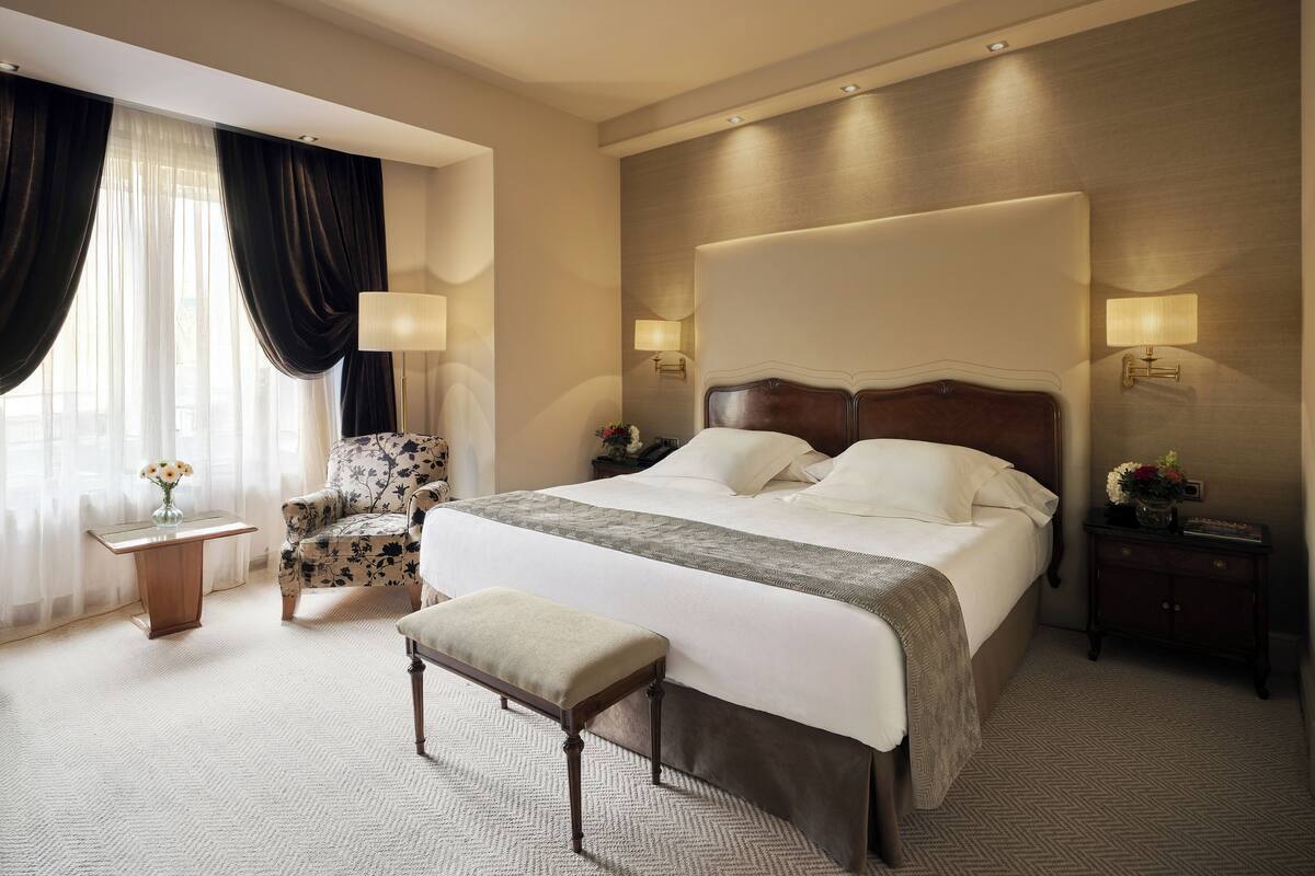 Quarto de hotel com uma cama grande, uma cadeira, um banquinho, uma mesa de cabeceira e um abajur.