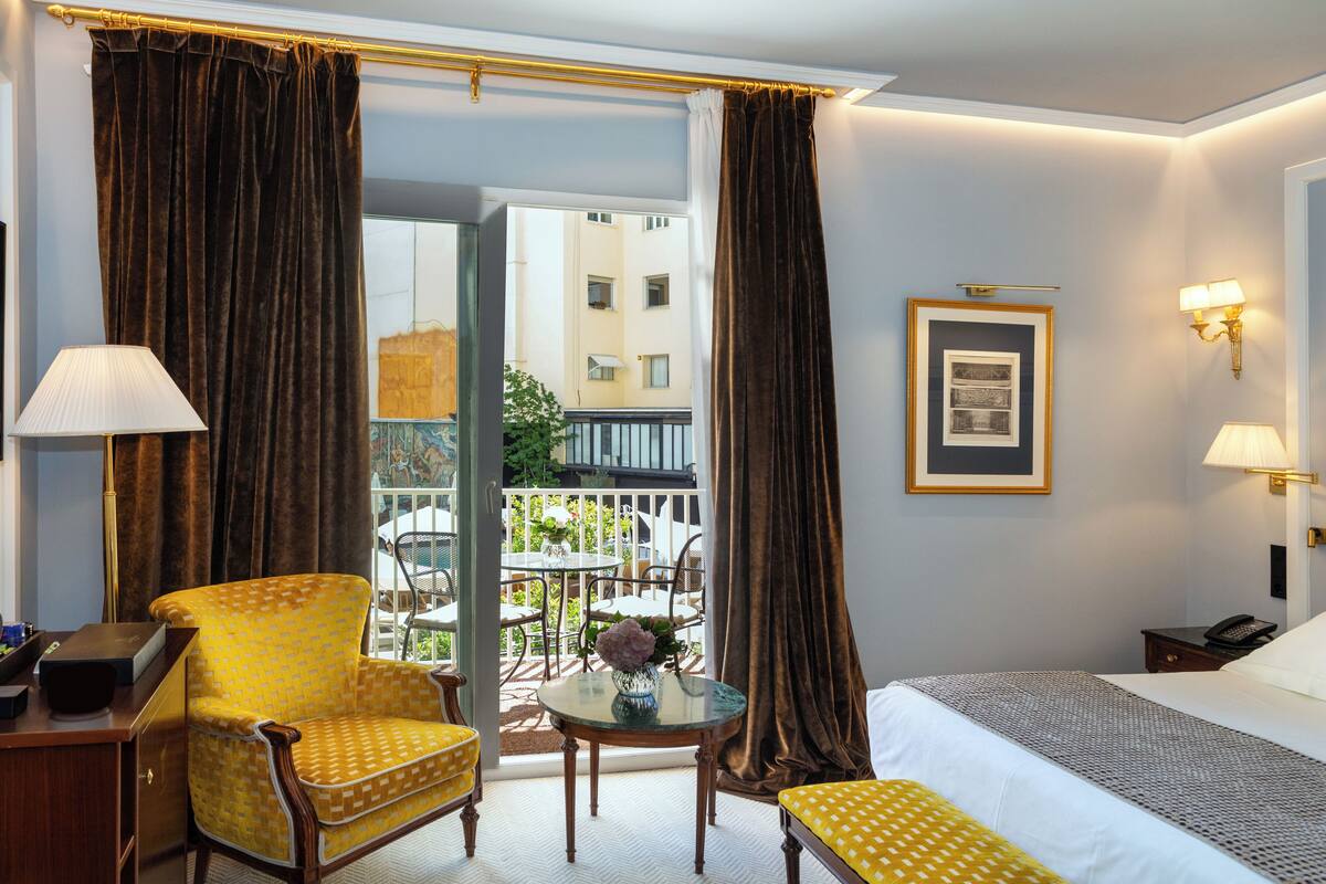 Quarto de hotel com cama, poltrona amarela, pequena mesa com um vaso e vista para a varanda externa.