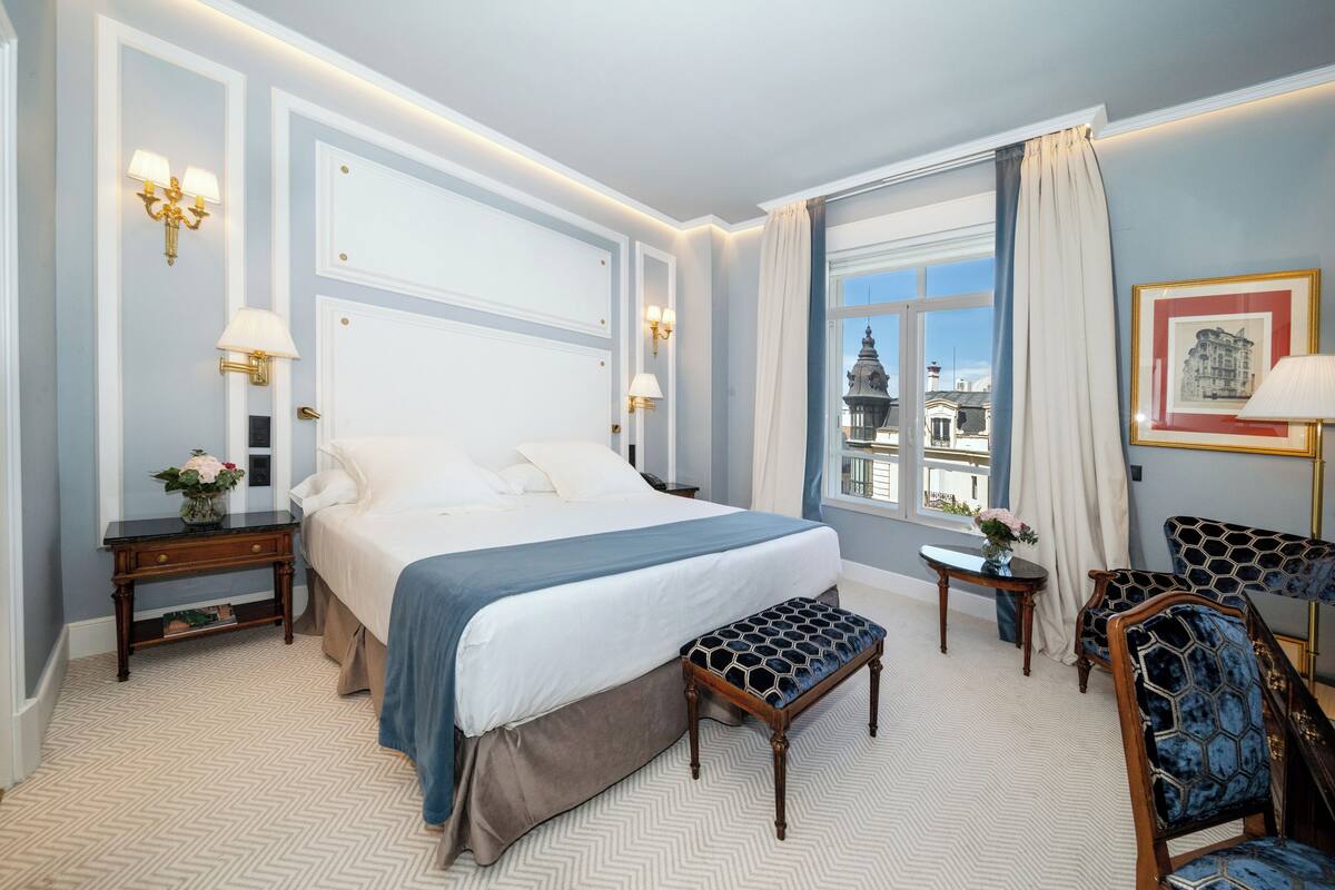 Quarto de hotel com uma cama grande, um banco, uma mesinha com um vaso e vista para o horizonte urbano através de uma janela.